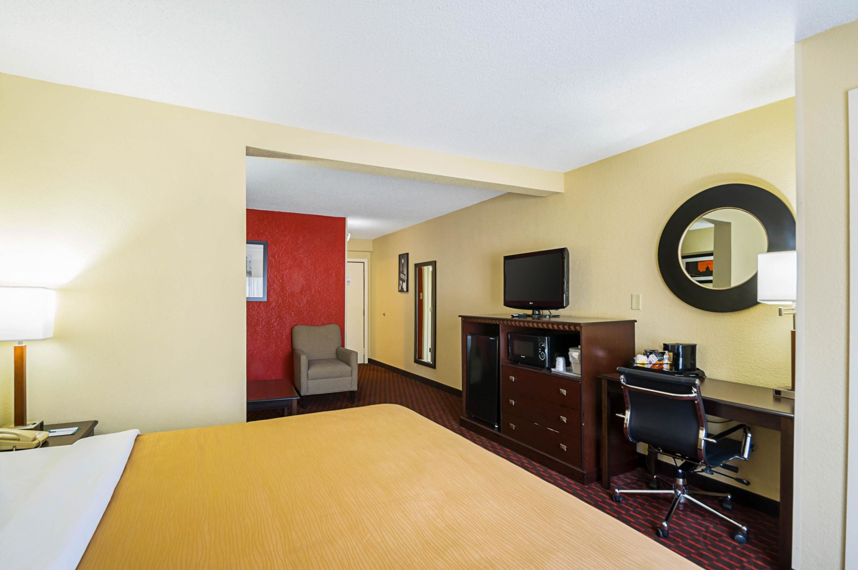 Quarto de hotel com cama, escrivaninha com computador, cadeira, televisão e espelho.