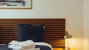Cama bem arrumada com travesseiro azul escuro, toalha branca e cabeceira de madeira. Uma mesinha com abajur ao lado da cama.