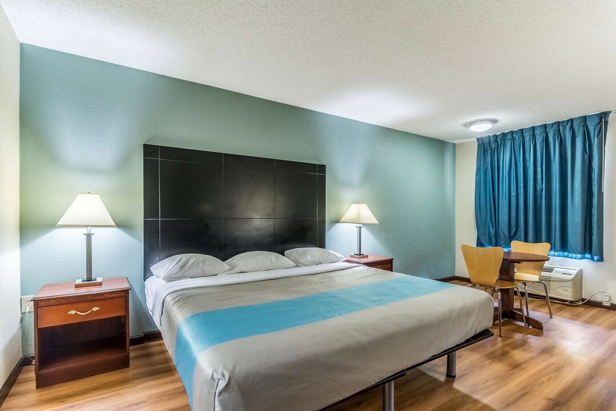 Quarto de hotel com cama, duas luminárias de cabeceira, uma mesa de cabeceira de madeira, uma escrivaninha com cadeira e uma janela com cortinas.