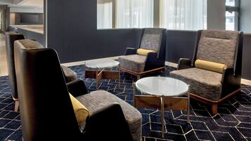 Um hall de hotel moderno com um tapete estampado em padrão geométrico, assentos confortáveis com almofadas e uma grande janela com vista para a cidade.