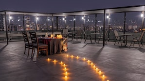 Terraço no telhado com uma mesa posta para dois, rodeado de cadeiras, e uma fileira de velas formando um coração no chão.