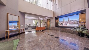 Um hall amplo com teto alto, uma grande janela com vista para a cidade, uma área de estar com um sofá e um banco, e uma planta em vaso.