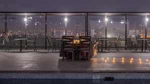 Terraço no telhado com mesas, cadeiras, piscina e luzes da cidade ao fundo.