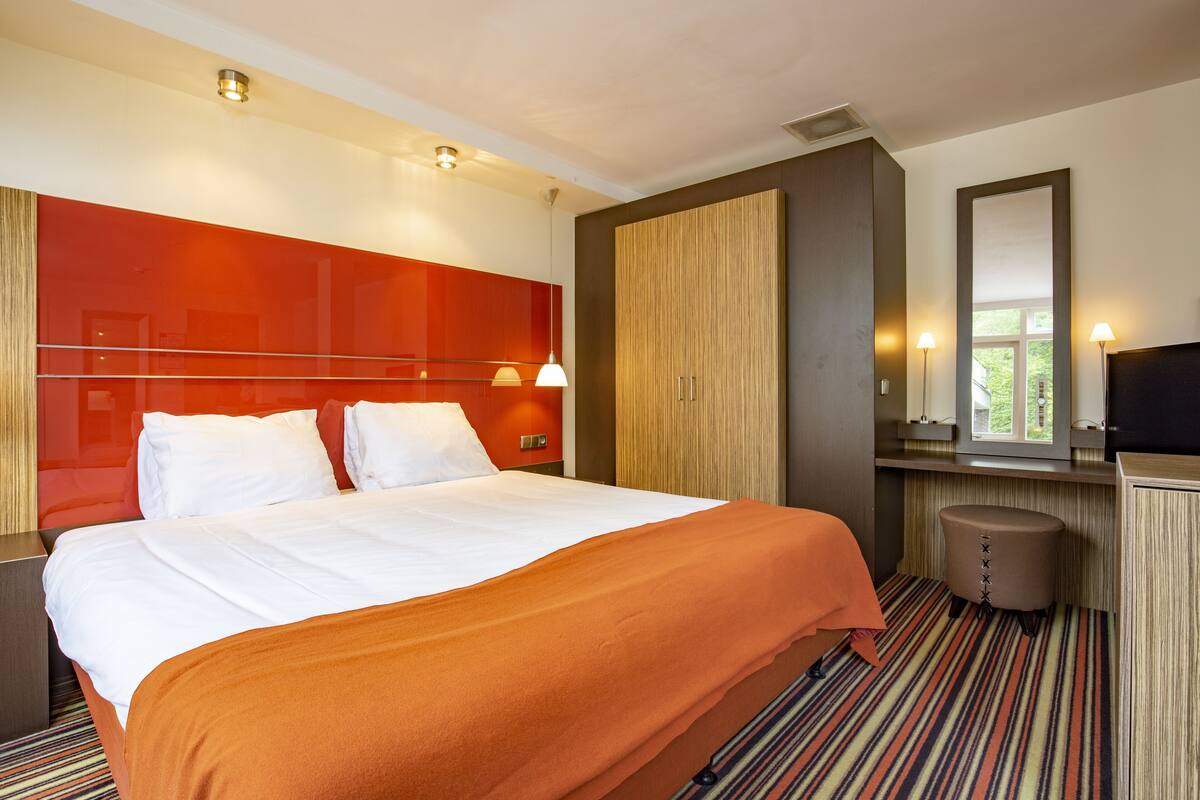 Quarto de hotel com uma cama grande, um guarda-roupa de madeira, um banquinho pequeno, uma televisão e uma janela com vista para o verde.