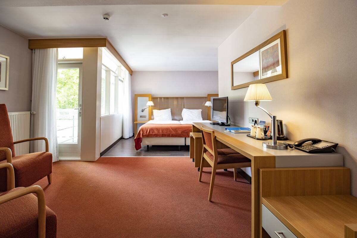 Quarto de hotel com cama, escrivaninha, cadeiras, abajur e telefone.