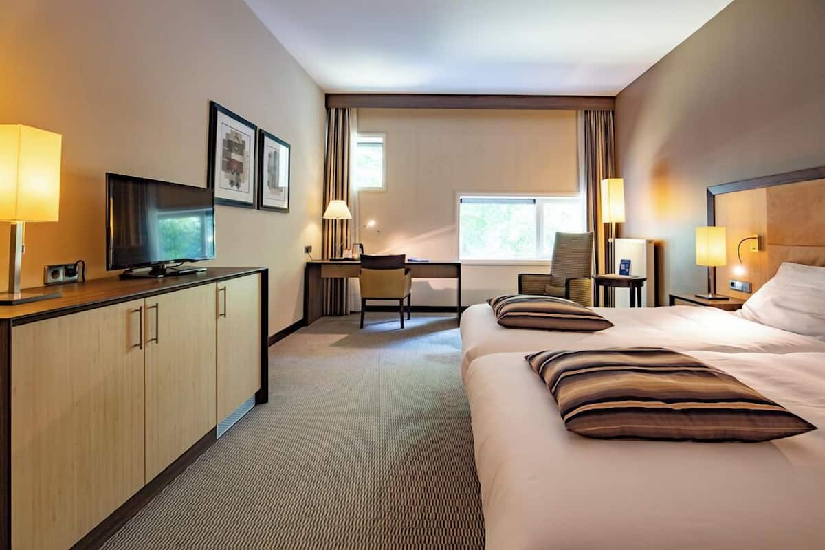 Quarto de hotel com cama, escrivaninha, cadeira, televisão e luminárias.