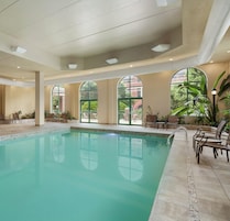 Piscina interior com paredes de vidro transparente, áreas de estar e plantas em vasos.