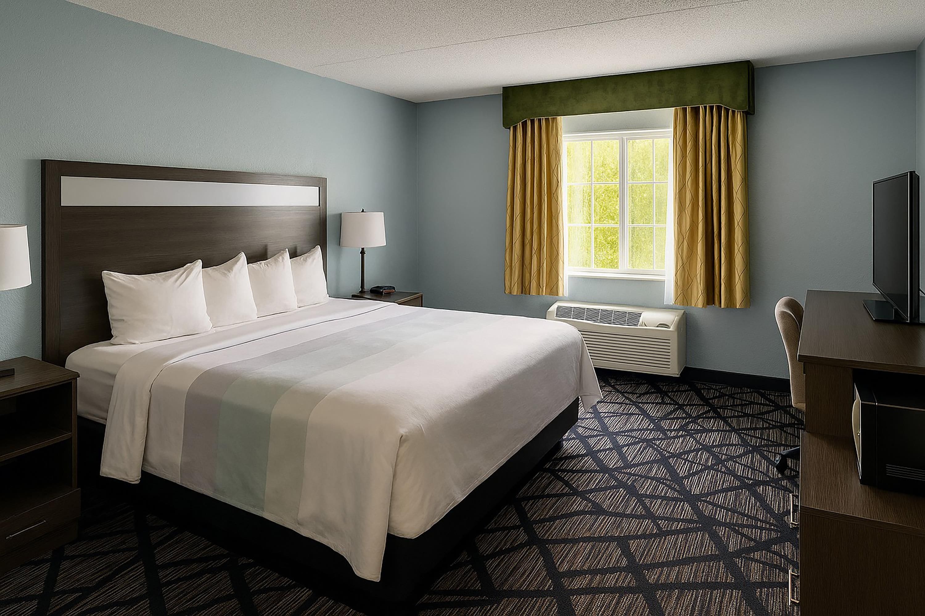 Quarto de hotel com uma cama grande, luminárias de cabeceira, janela com cortinas, escrivaninha com televisão de tela plana e ar-condicionado fixado na parede.