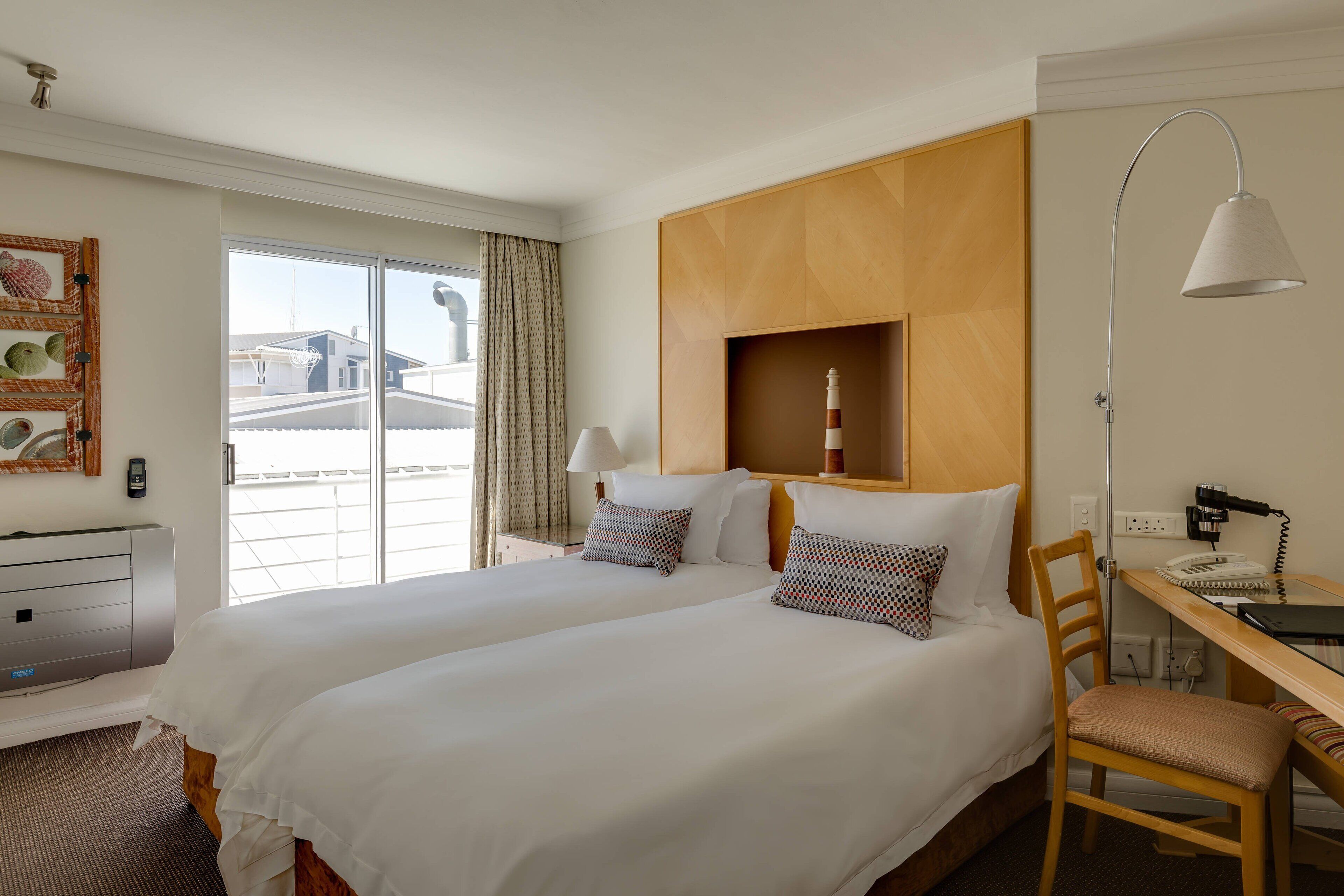 Quarto de hotel com duas camas, uma escrivaninha, uma cadeira, um abajur e vista para o exterior.