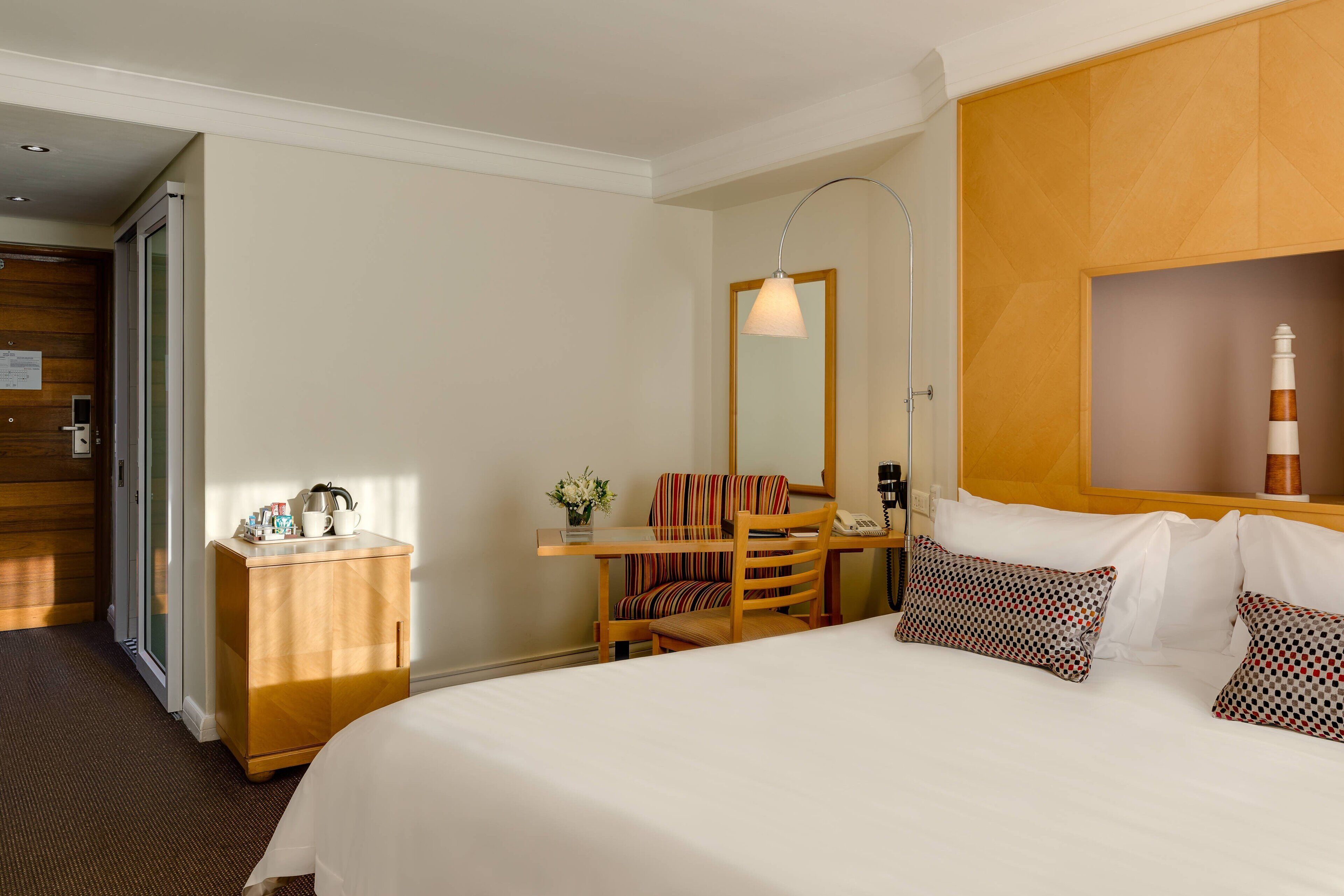 Quarto de hotel com cama, escrivaninha com cadeira, espelho e guarda-roupa de madeira.
