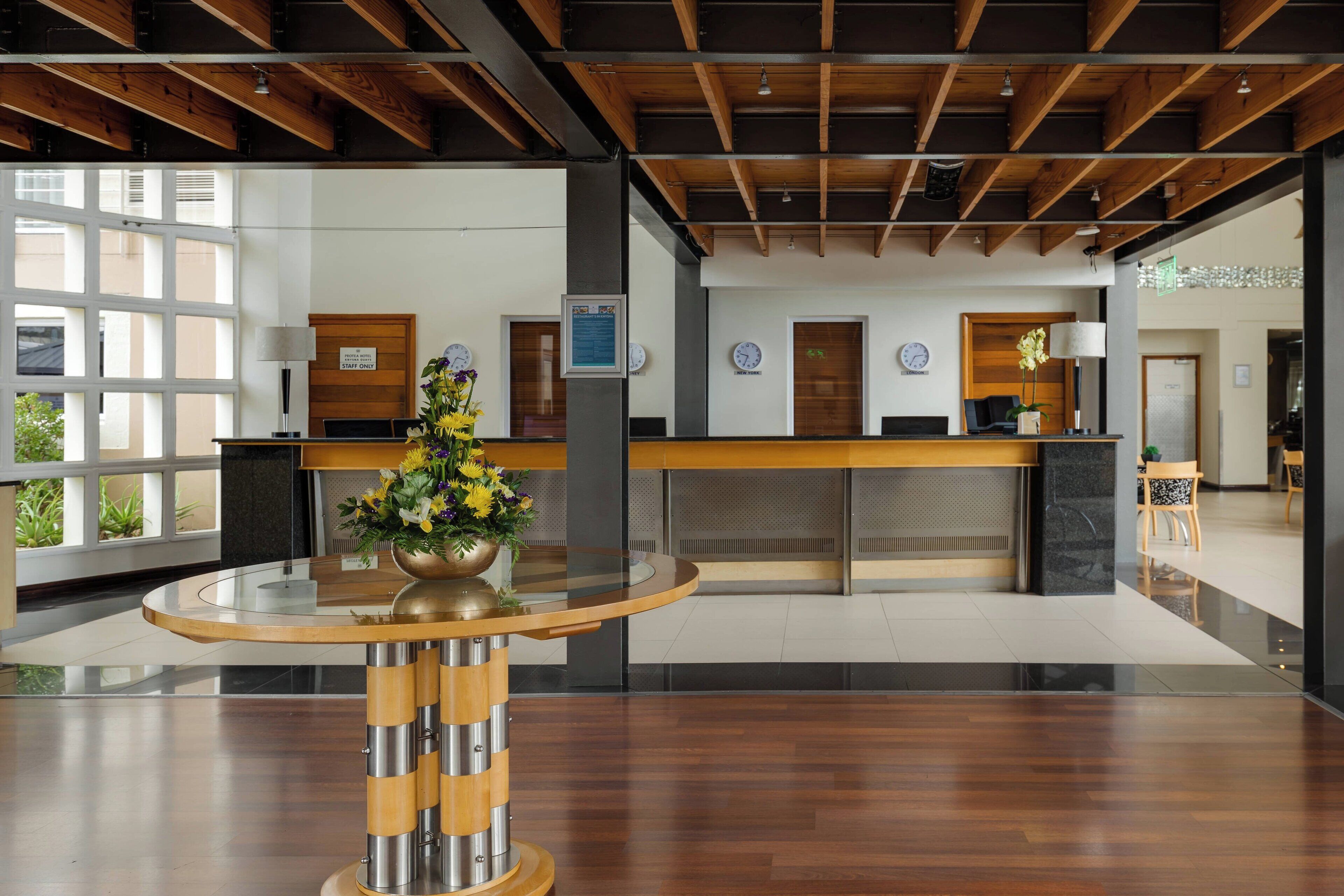 Uma área de recepção com balcão de madeira, uma mesa redonda com um vaso de flores e design interior moderno com vigas de madeira e painéis de vidro.