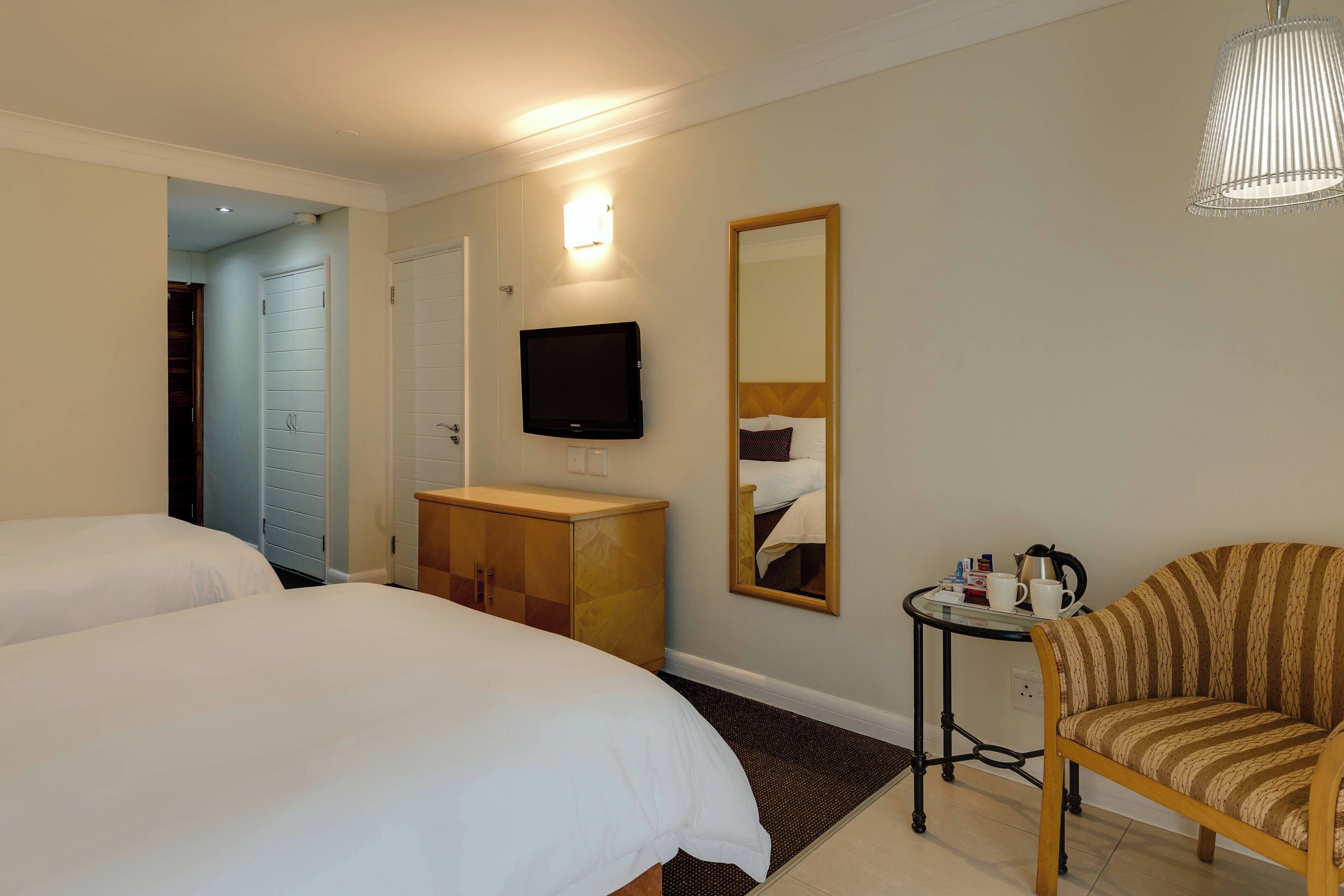 Quarto de hotel com duas camas, televisão, espelho, mesinha com uma cadeira e uma luminária.
