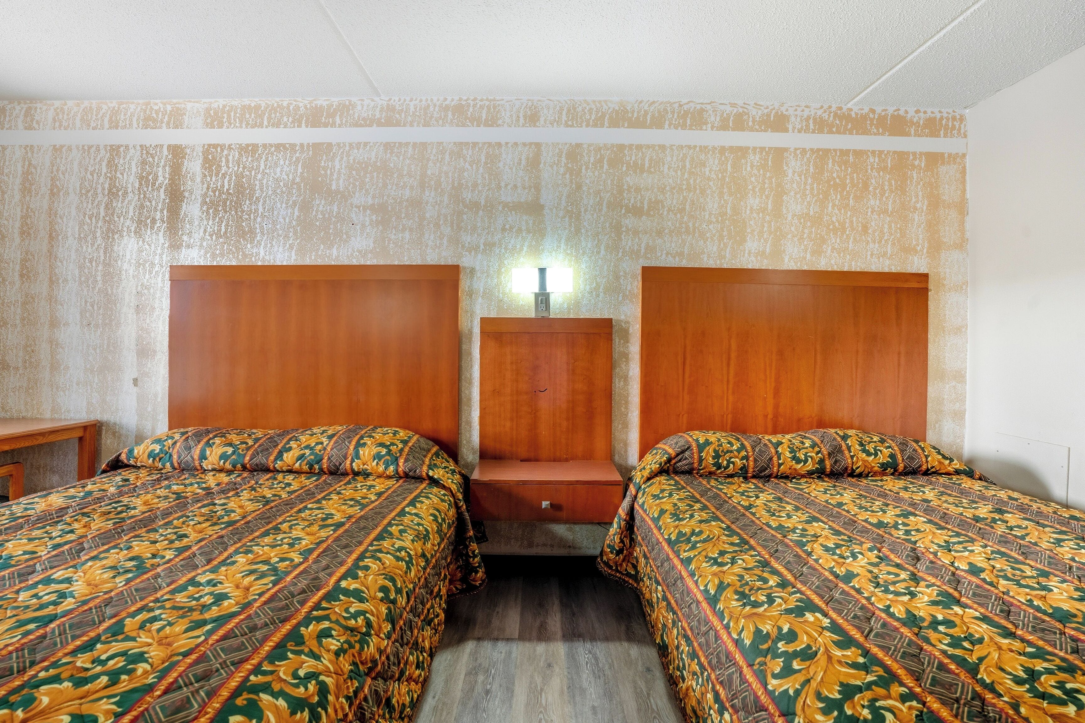 Quarto com duas camas de solteiro, cabeceiras de madeira, mesa de cabeceira e luminária fixa na parede.