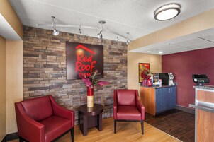 Área de descanso Red Roof com duas cadeiras vermelhas, uma mesa pequena com um vaso, uma parede de tijolos, um balcão de recepção e uma máquina de café.