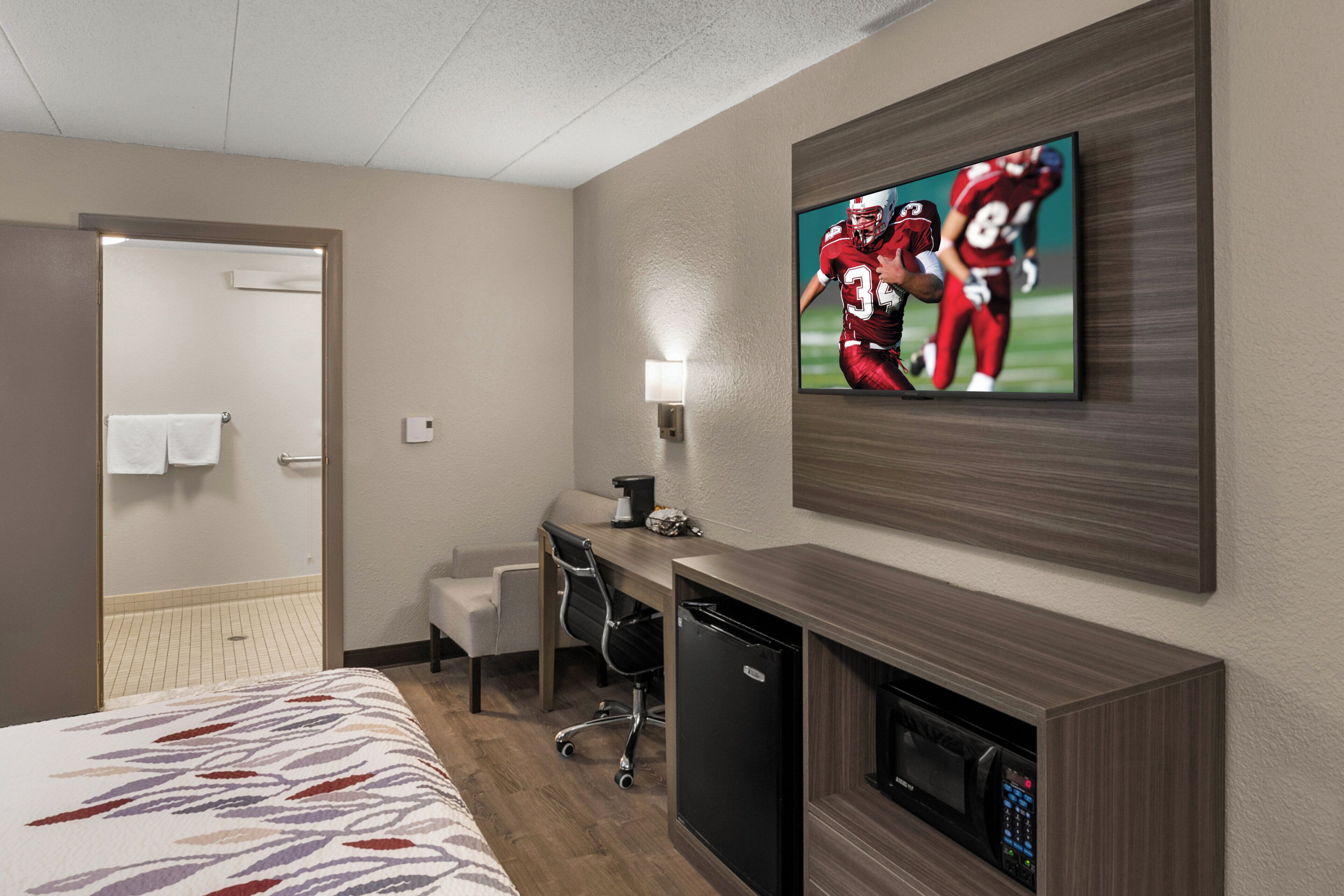 Quarto de hotel com cama, escrivaninha com cadeira, televisão na parede e micro-ondas.