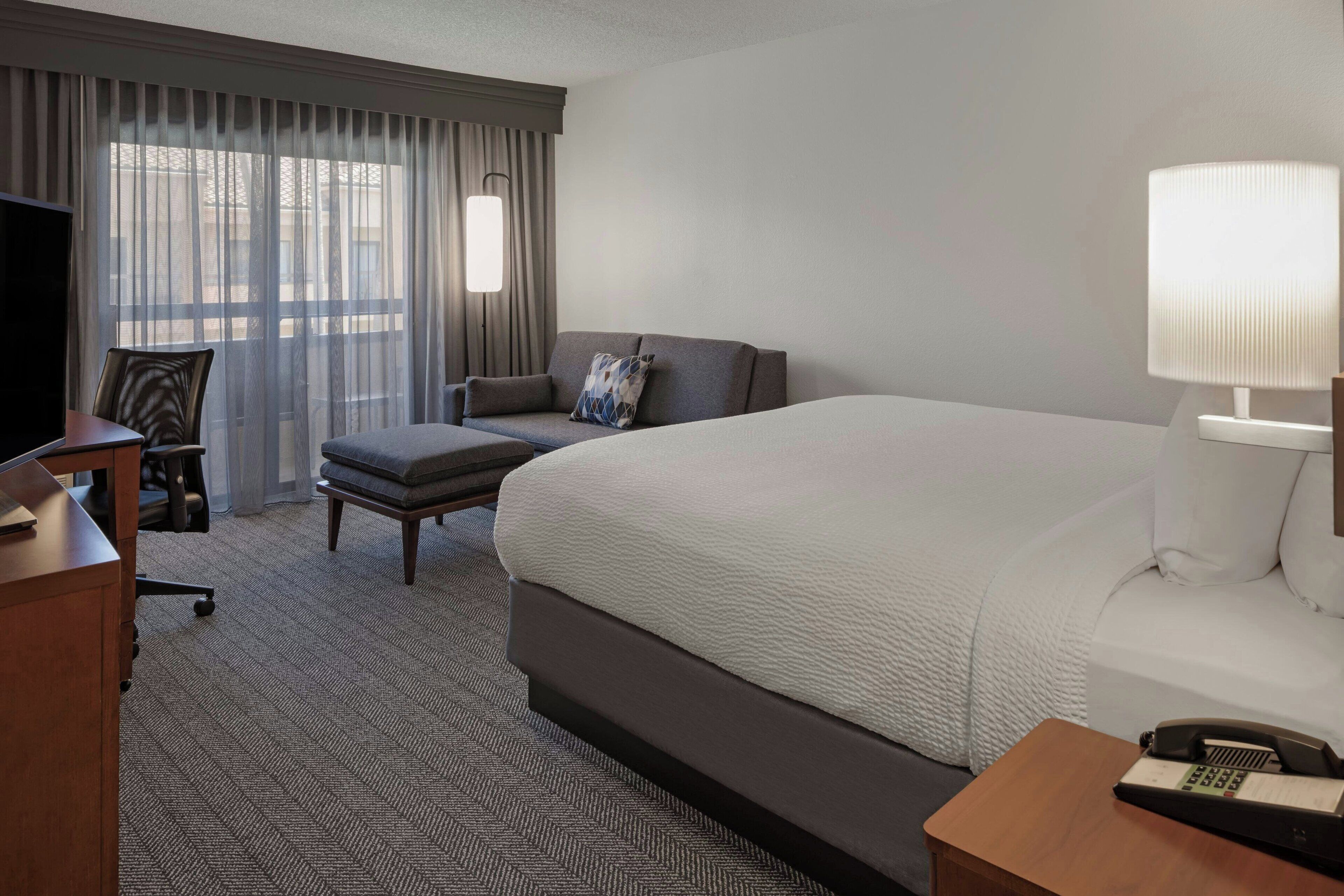 Quarto de hotel com uma cama grande, uma escrivaninha com telefone, uma cadeira e uma janela com cortinas.