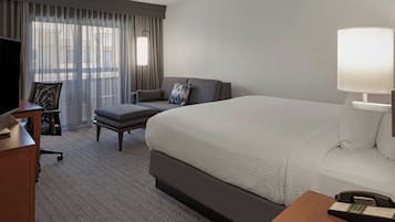 Quarto de hotel com uma cama grande, uma escrivaninha com telefone, uma cadeira e uma janela com cortinas.