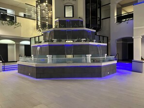 Uma cascata escalonada com laterais de vidro e iluminação azul, localizada em um amplo espaço interno com teto alto e arquitetura moderna.