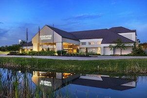 A hotel with a modern design, featuring a large sign for "HOMEWOOD SUITES by Hilton" and a reflection in a pond in front.