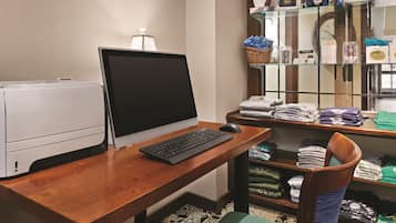 Uma mesa de madeira com um laptop, teclado e rato. Uma impressora está à esquerda. Atrás da mesa, há prateleiras com roupas dobradas e uma cesta de vime.