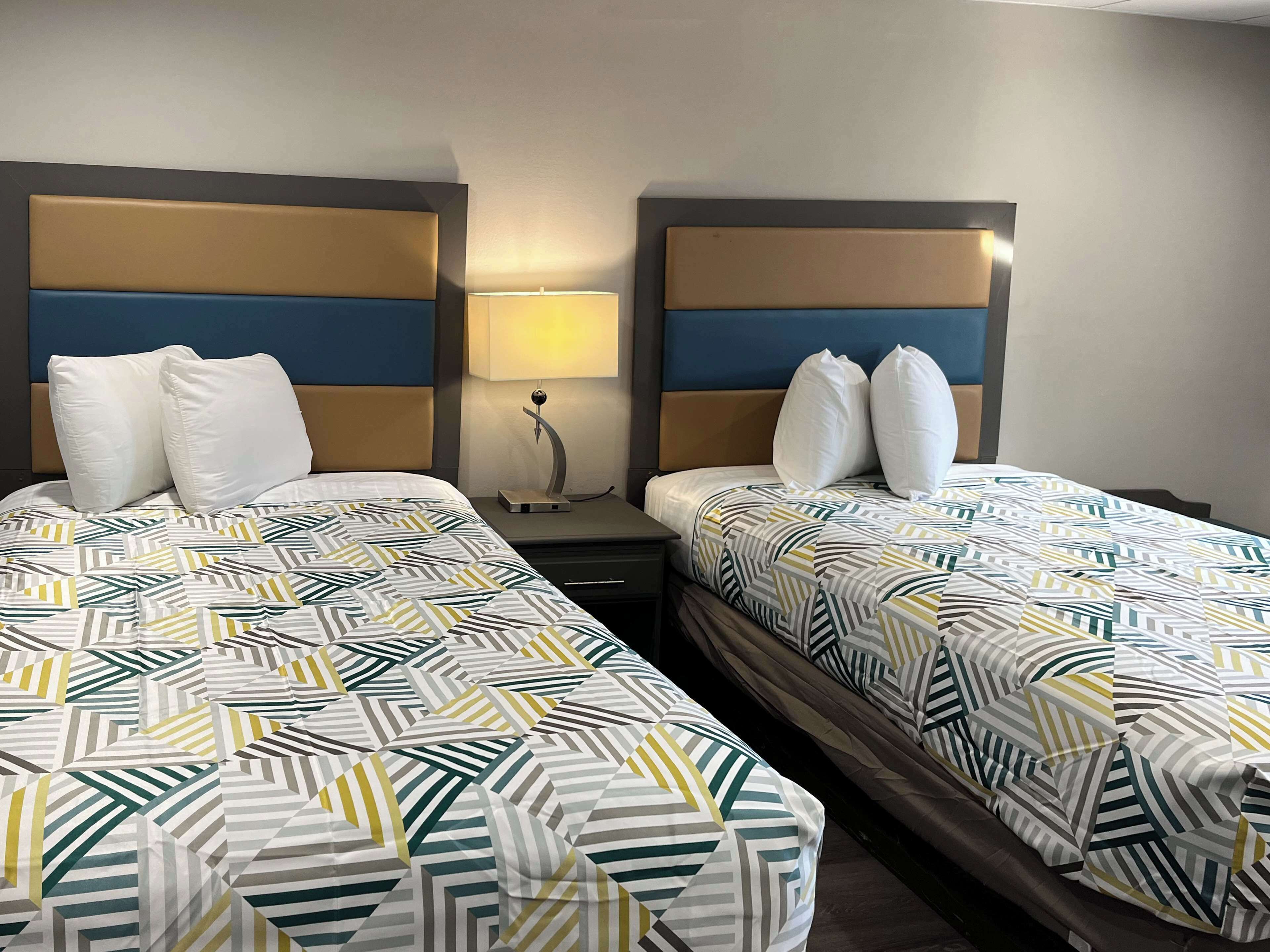 Quarto de hotel com duas camas, cada uma com colcha estampada e almofadas brancas. Há uma mesa de cabeceira com abajur entre as camas.