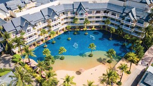 Vista aérea de um resort com uma piscina central rodeada por palmeiras, várias varandas e uma área de praia.