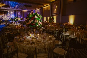 Salão de festas com mesas redondas preparadas para um evento formal, com centros de mesa florais e elegantes louças.