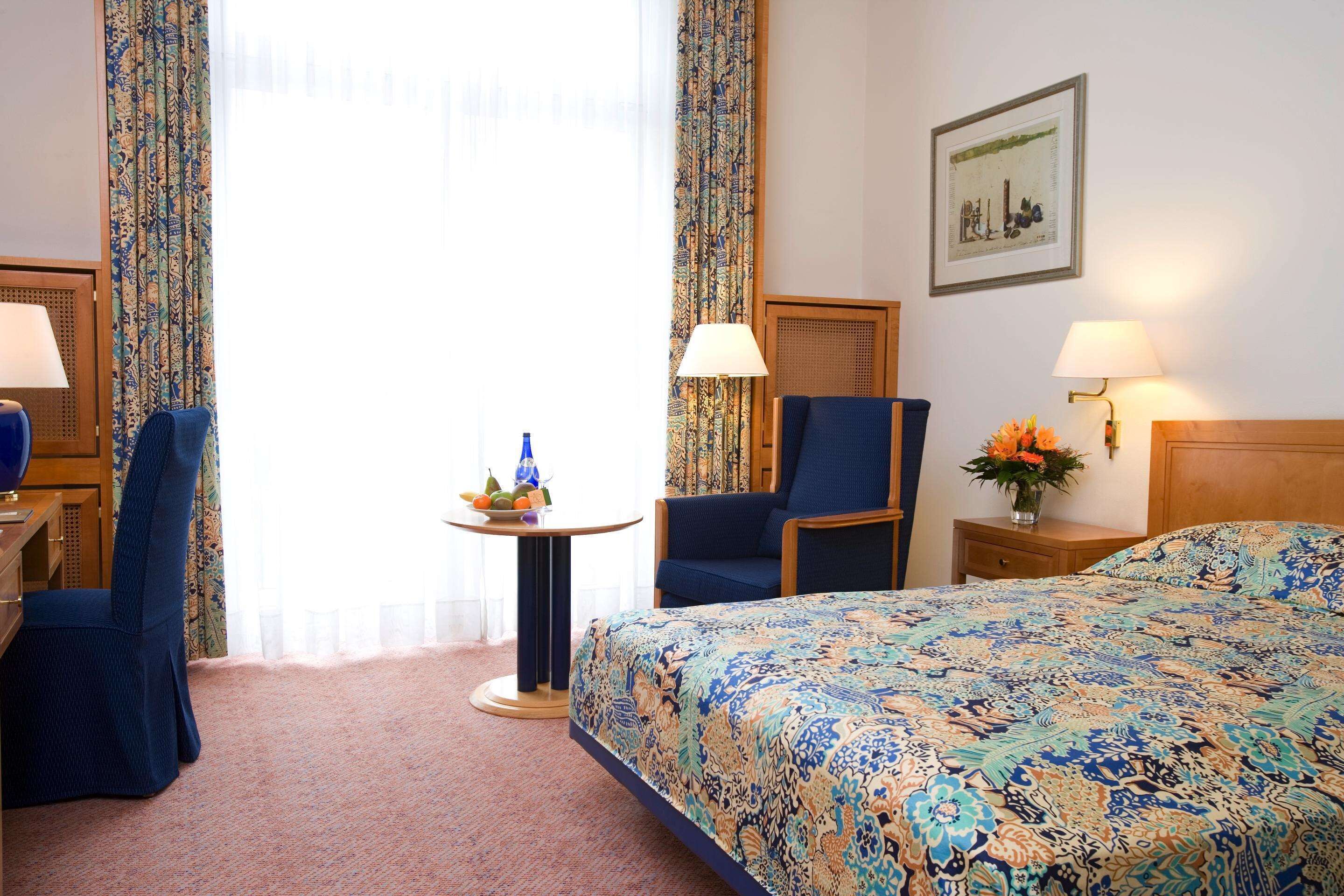 Quarto de hotel com cama, duas cadeiras, uma mesinha com abajur e um quadro na parede.