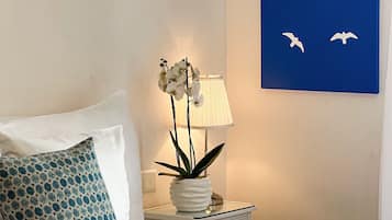Quarto de hotel com cama, mesa de cabeceira com luminária, orquídea em vaso e painel azul com aves brancas.