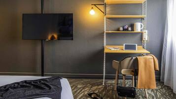 Quarto de hotel com cama, escrivaninha com laptop, televisão e prateleira com objetos.