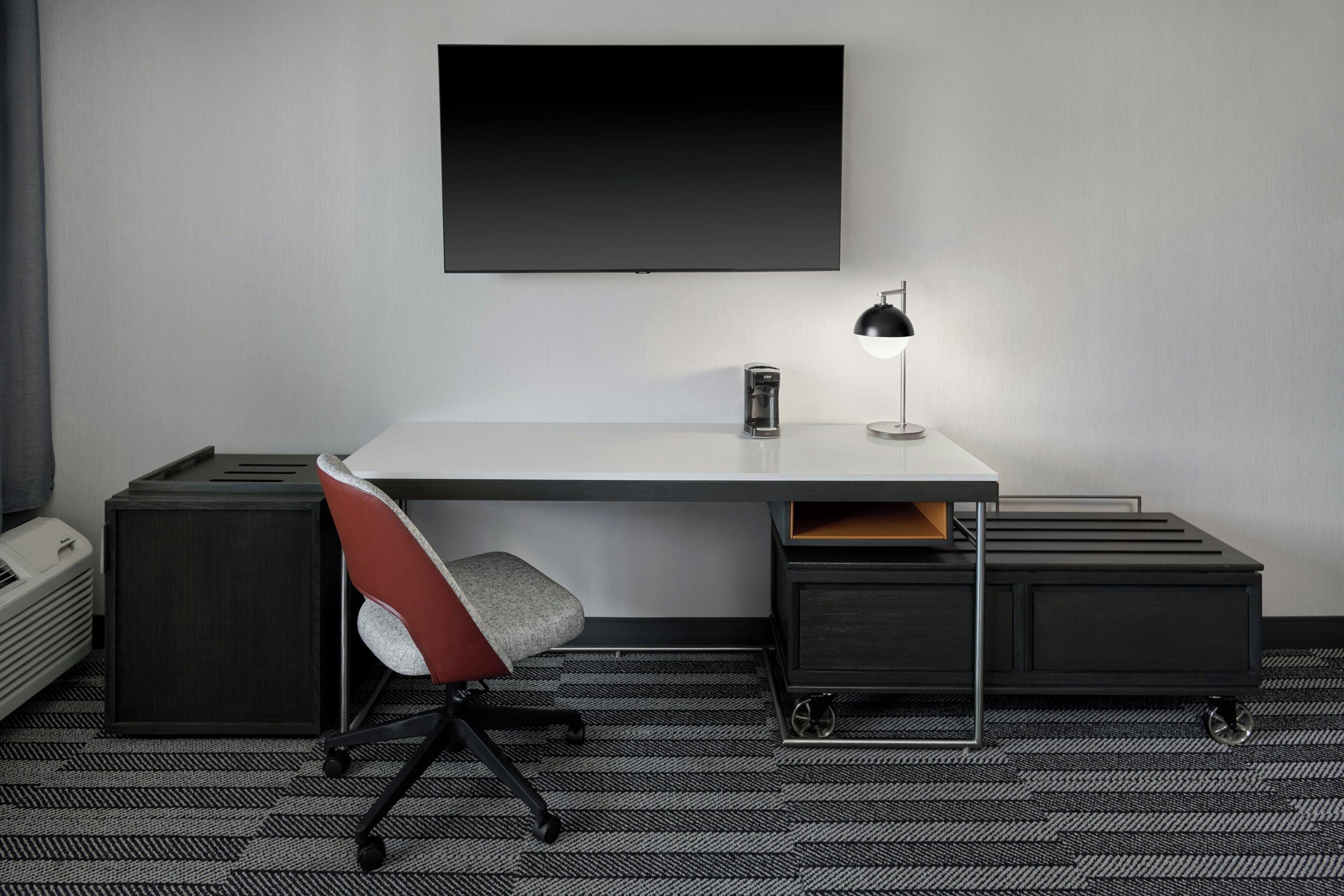 Um espaço de escritório moderno com uma mesa branca, uma luminária preta, uma cadeira vermelha e cinza, e uma TV de tela plana fixada na parede.