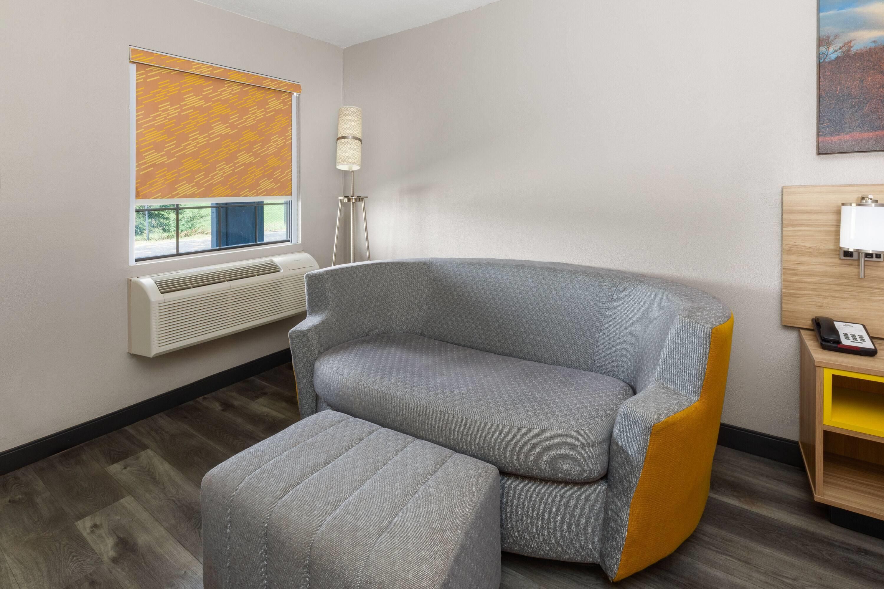 Quarto de hotel com poltrona cinza, um banquinho do mesmo tom, luminária de chão, mesa de cabeceira com telefone e janela com persianas.