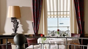 Uma área de refeições com vista para o mar, mesa posta com toalhas brancas e rosas vermelhas, e um abajur à esquerda.