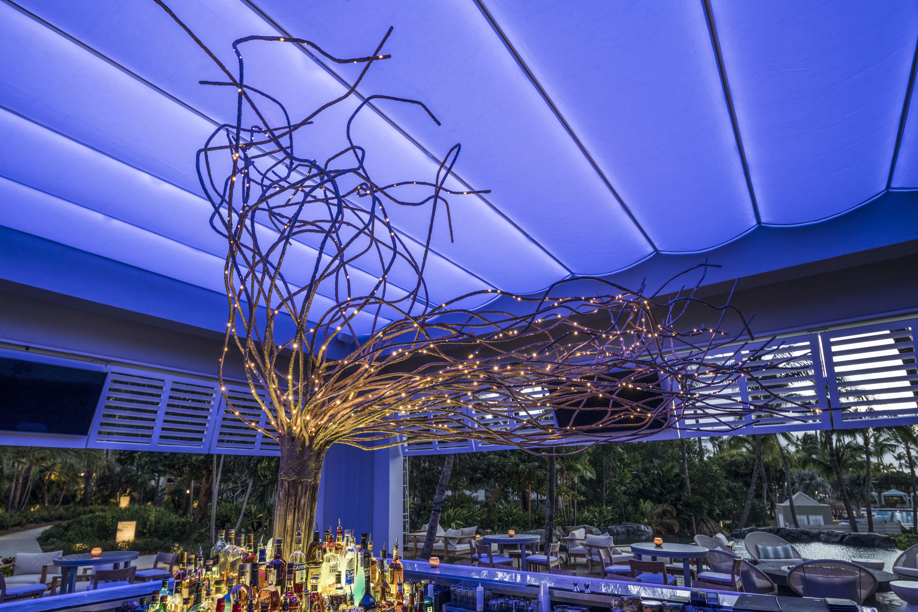 Uma área de bar com uma instalação de iluminação incomum em formato de árvore, mesas ao ar livre e vista para a piscina.