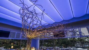 Uma área de bar com uma instalação de iluminação incomum em formato de árvore, mesas ao ar livre e vista para a piscina.