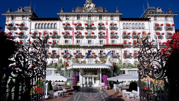 Um hotel imponente com várias varandas adornadas com flores vermelhas, uma entrada central com marquise decorativa e áreas de estar ao ar livre.