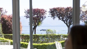 Uma área de refeições com mesa e cadeiras, um vaso com flores e vista para o mar através de grandes janelas.