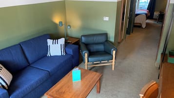 Quarto com sofá azul, mesa de madeira, poltrona de couro preto e uma cama ao fundo.