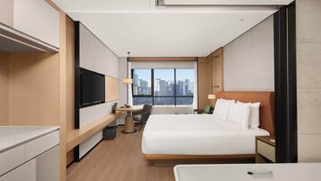 Quarto de hotel moderno com uma cama grande, uma escrivaninha com cadeira, televisão e janela com vista para a cidade.