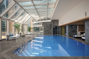 Piscina interior com teto de vidro, assentos modernos e chão em mosaico de pedra.