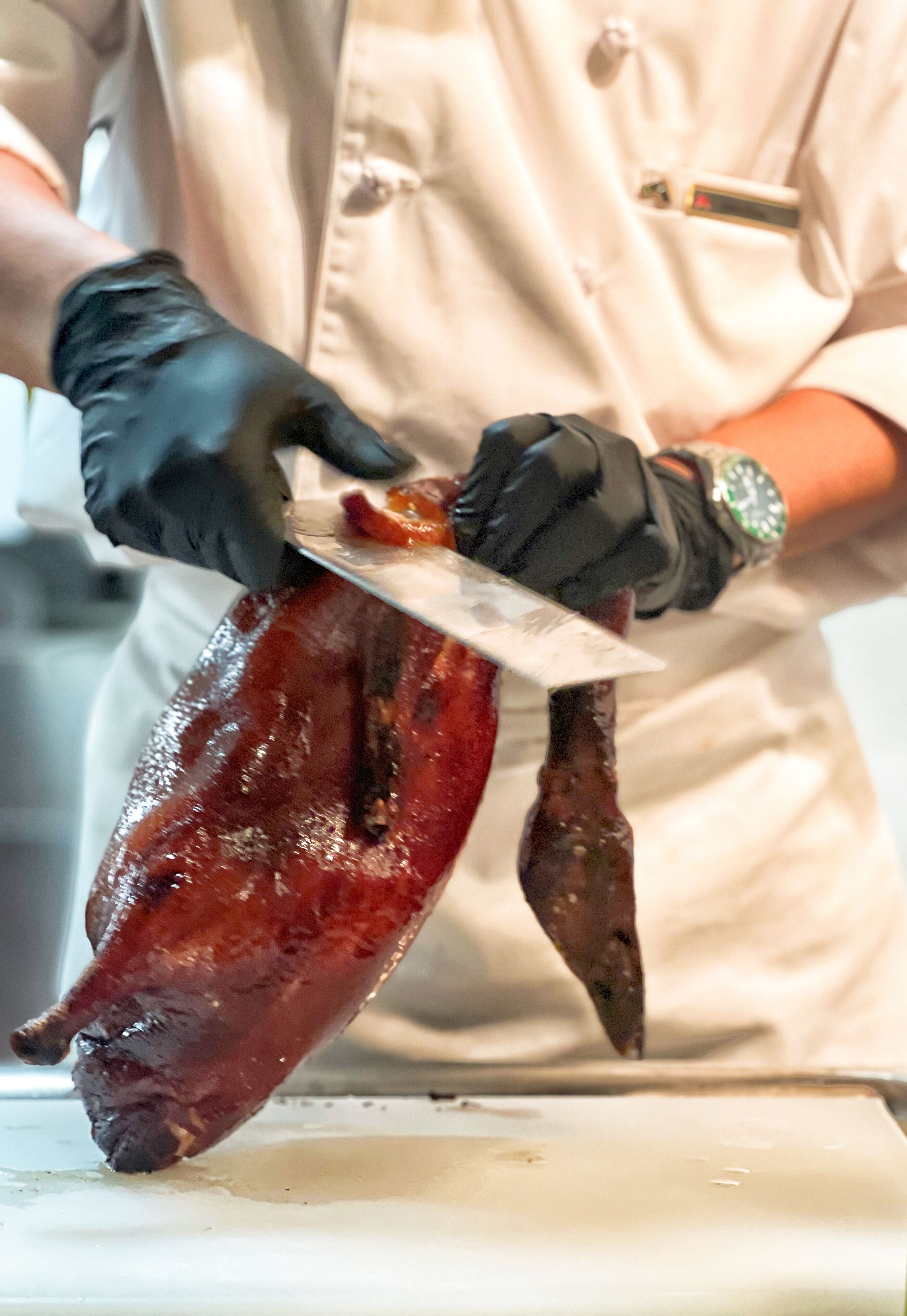 Uma pessoa com avental branco e luvas pretas segurando um pedaço grande de carne vermelha e uma faca.