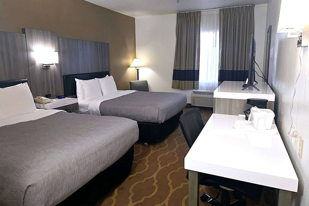 Quarto de hotel com duas camas, uma mesa, uma cadeira, televisão, luminária e janela com cortinas.