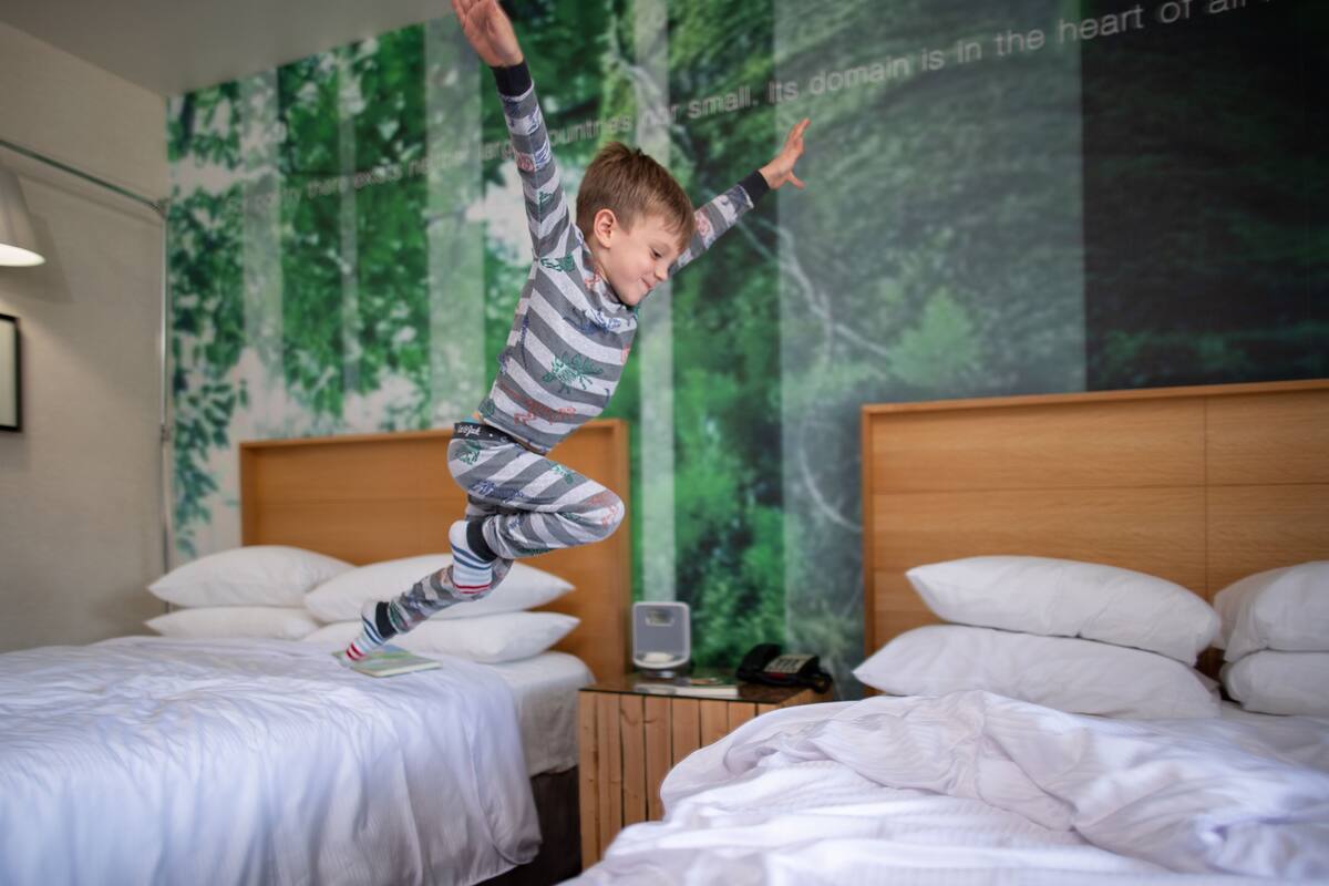 Criança pulando em uma cama em quarto de hotel com um grande mural temático de floresta.