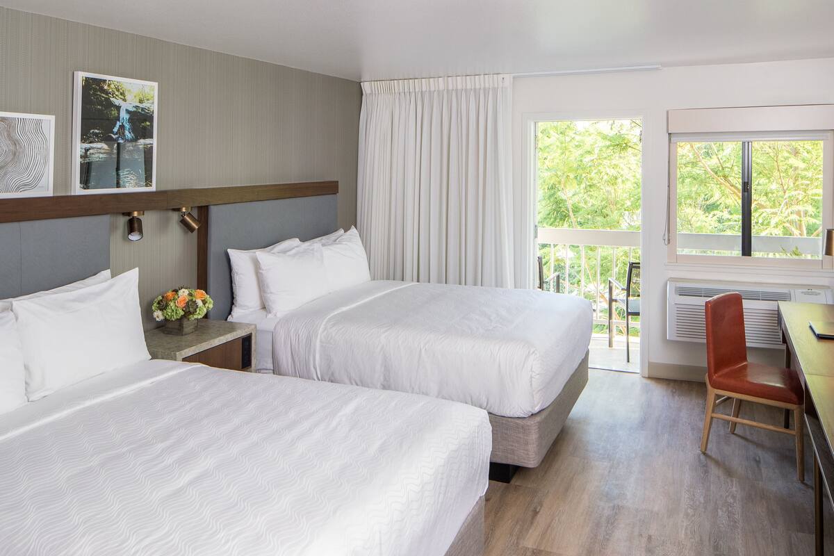Quarto de hotel com duas camas, uma escrivaninha, uma cadeira, janela com cortinas e obras de arte na parede.