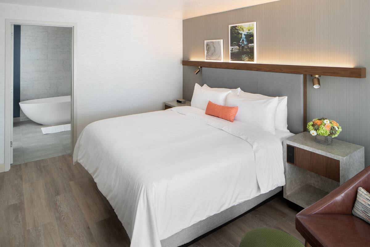 Quarto de hotel moderno com uma cama grande, uma mesinha de cabeceira com abajur, uma pequena mesa com um vaso de flores e vista para um banheiro com banheira.