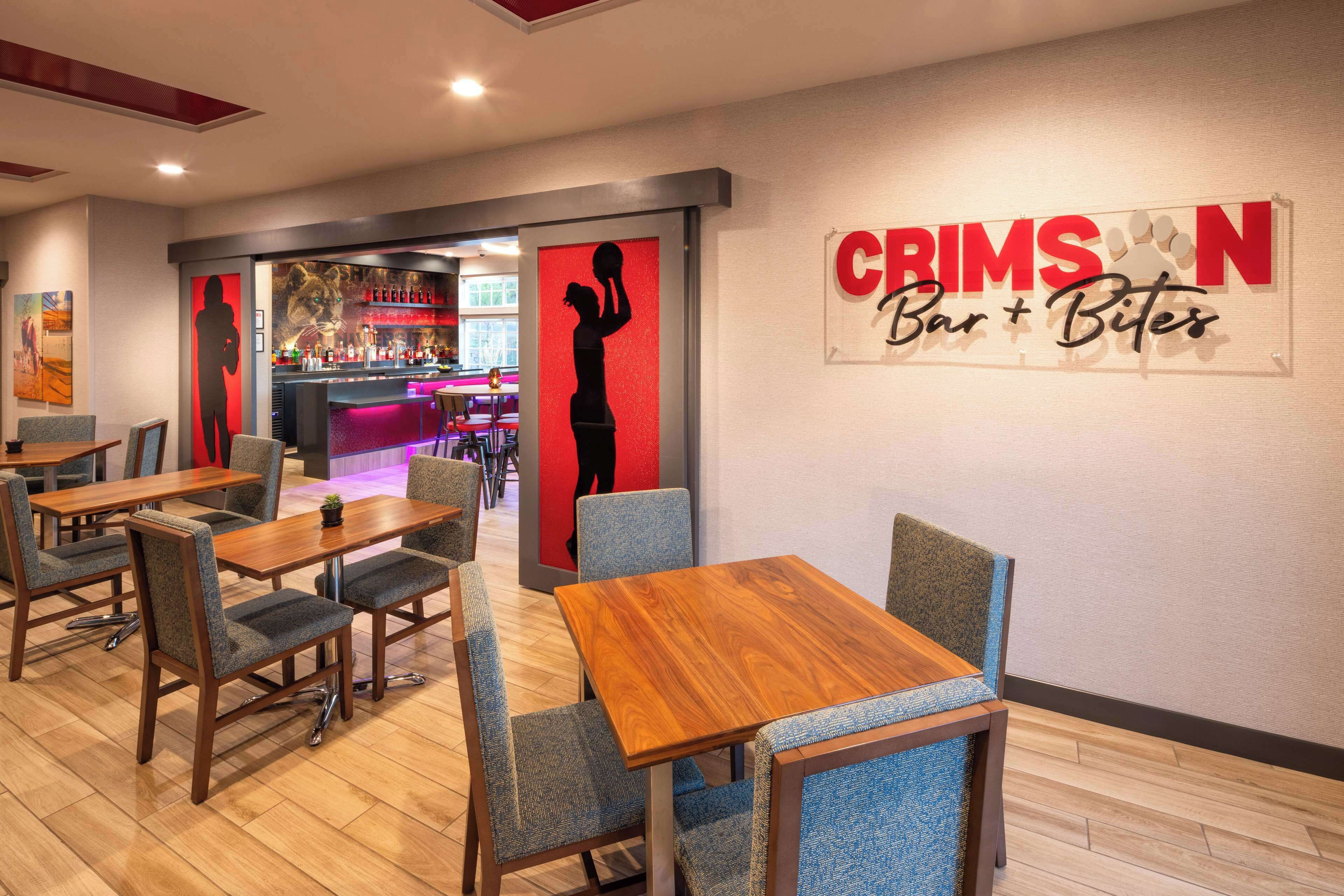 Interior de restaurante com mesas de madeira e cadeiras azuis, área de bar e placa com a inscrição 'CRIMSON Bar + Bites'.