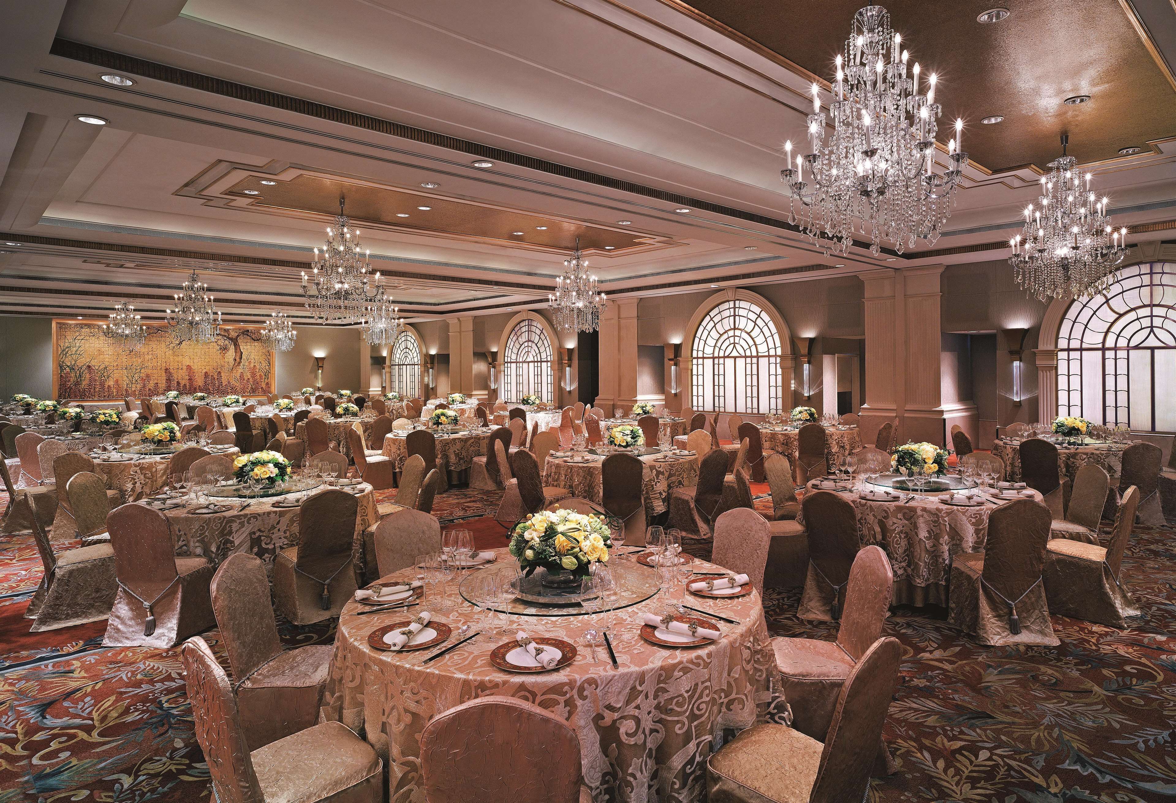 Uma sala de banquetes com mesas redondas, cadeiras, lustres e arranjos florais.