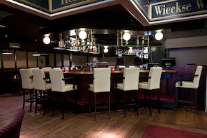Uma área de bar com balcão de madeira, banquetas brancas e diversos frascos e copos nas prateleiras.