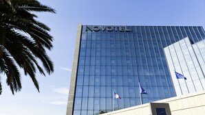 Edifício do hotel Novotel com fachada de vidro moderna, bandeiras da União Europeia e palmeiras em primeiro plano.