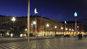 Uma praça com piso xadrez, postes de iluminação e edifícios ao fundo.